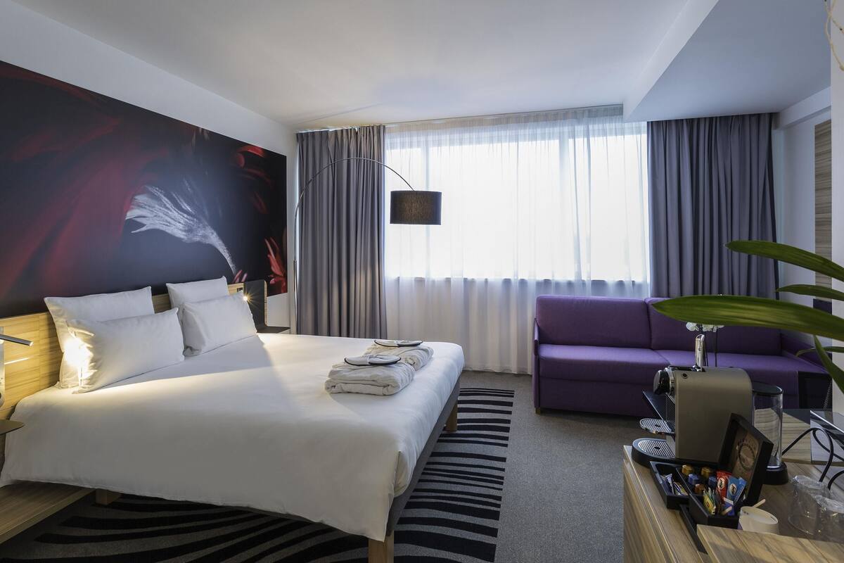 Quarto de hotel com cama, sofá, máquina de café e uma janela grande com cortinas.