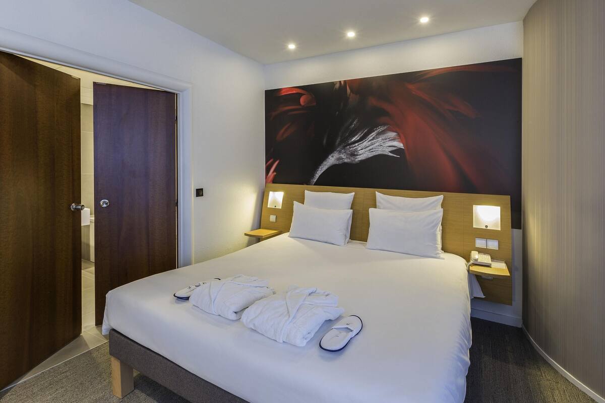 Quarto de hotel com uma cama grande, duas mesas de cabeceira, uma obra de arte pendurada na parede e uma porta.