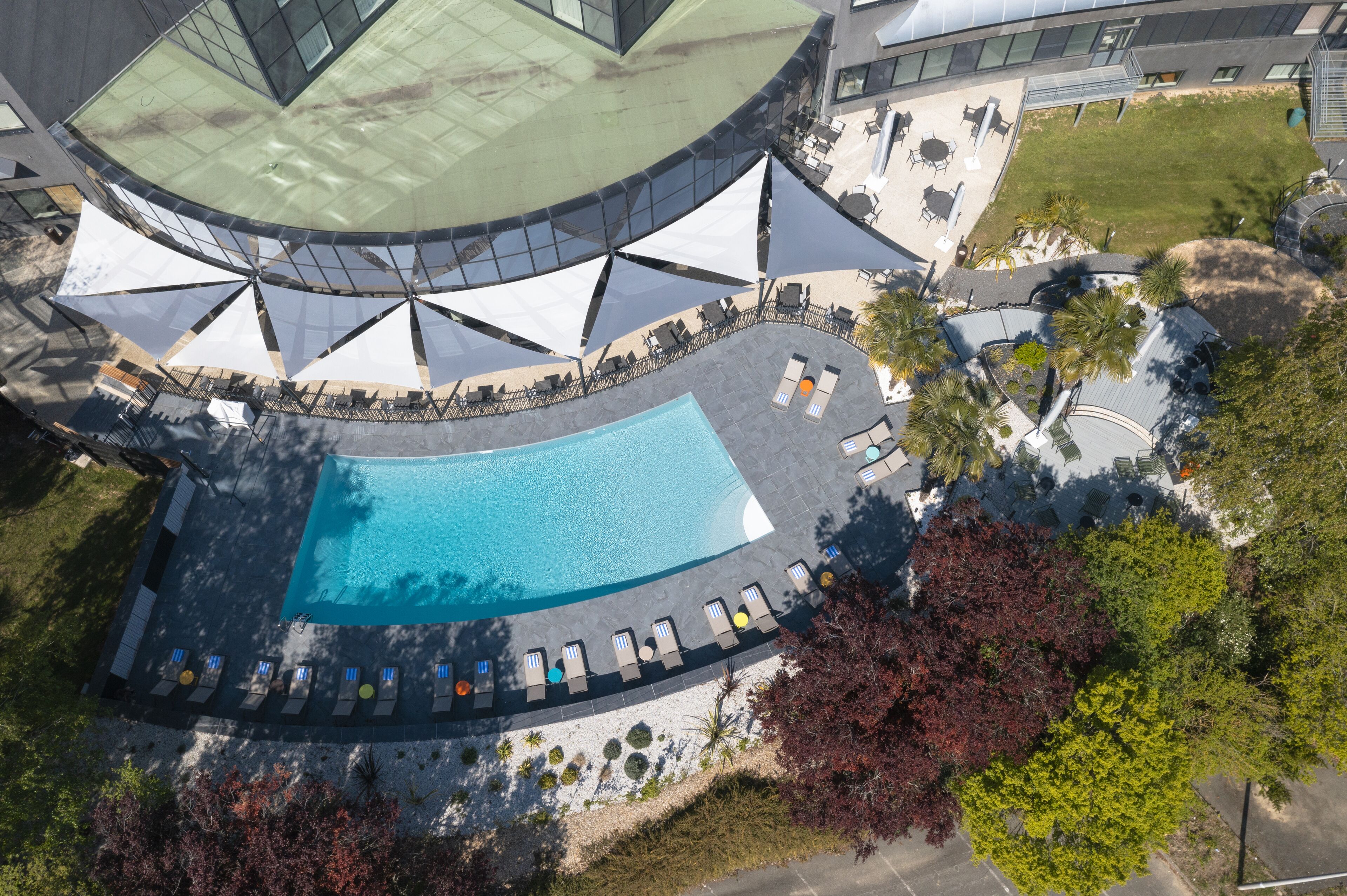 Vista aérea de um hotel com piscina, espreguiçadeiras e área externa coberta com guarda-sóis.