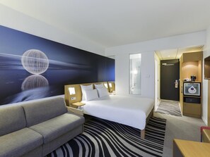 Quarto de hotel moderno com uma cama grande, um sofá, uma escrivaninha e um mural na parede.