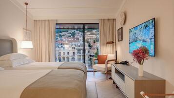 Quarto de hotel com duas camas, uma escrivaninha, uma cadeira, uma janela grande com vista para a cidade e um quadro na parede.