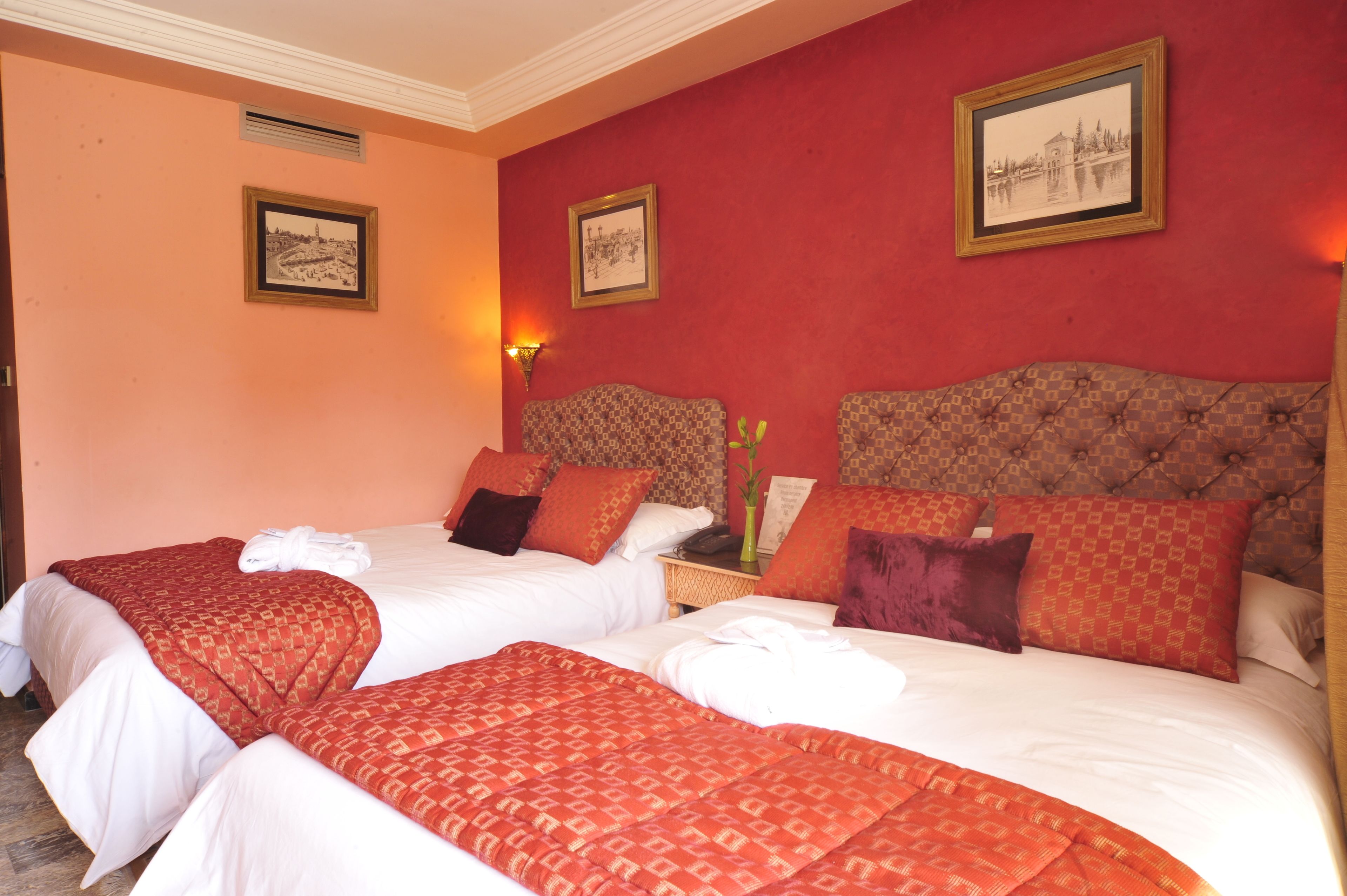 Quarto de hotel com duas camas, roupa de cama vermelha e branca, e duas molduras com imagens nas paredes.
