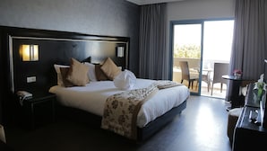 Quarto de hotel com uma cama grande, mesa de cabeceira com luminária, cadeira, pequena mesa com um vaso e vista para o exterior por uma porta deslizante.