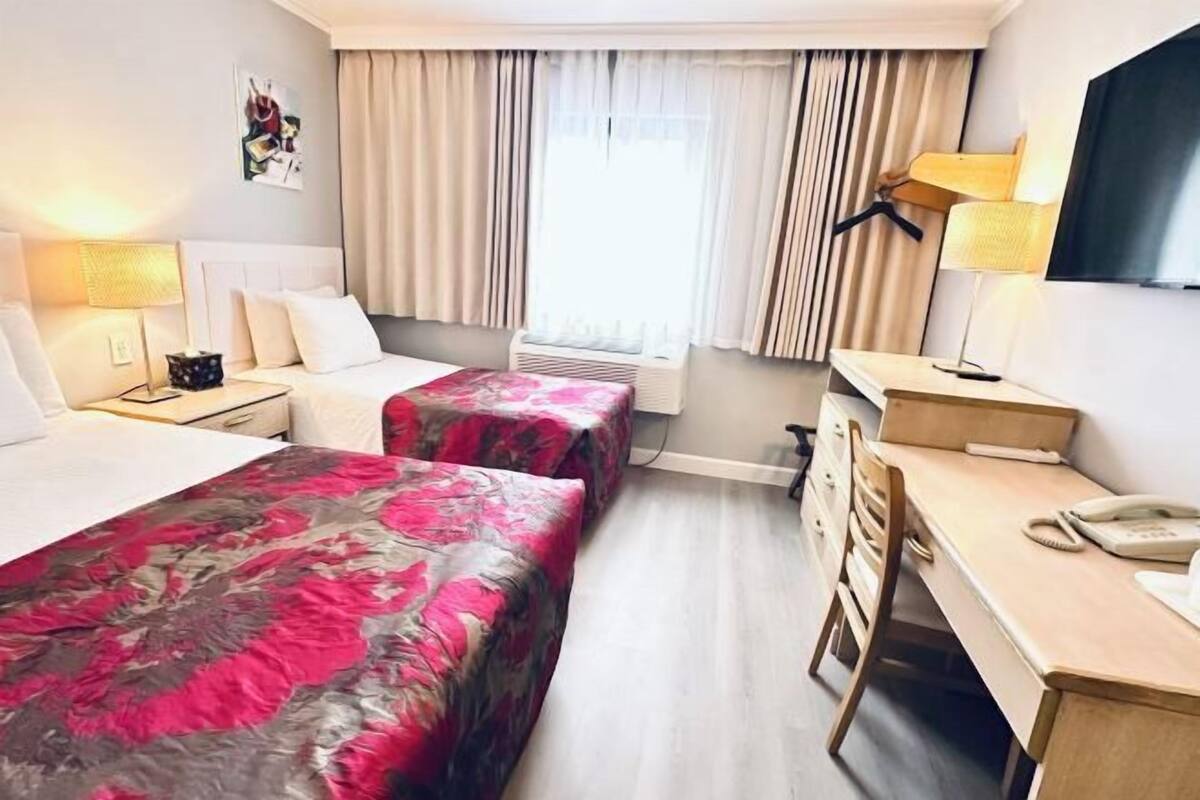 Quarto de hotel com duas camas, uma escrivaninha, uma cadeira, uma televisão e uma janela com cortinas.