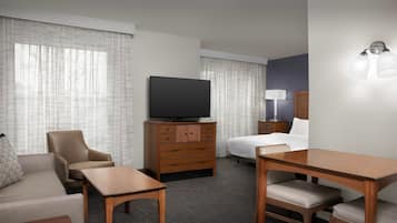 Quarto de hotel com sofá, mesa de madeira, televisão sobre um móvel de madeira, cama e mesa de cabeceira com abajur.