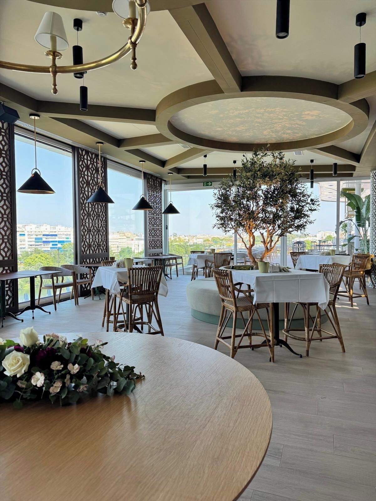 Restaurante com mesas redondas, cadeiras de vime e uma grande árvore ao centro.