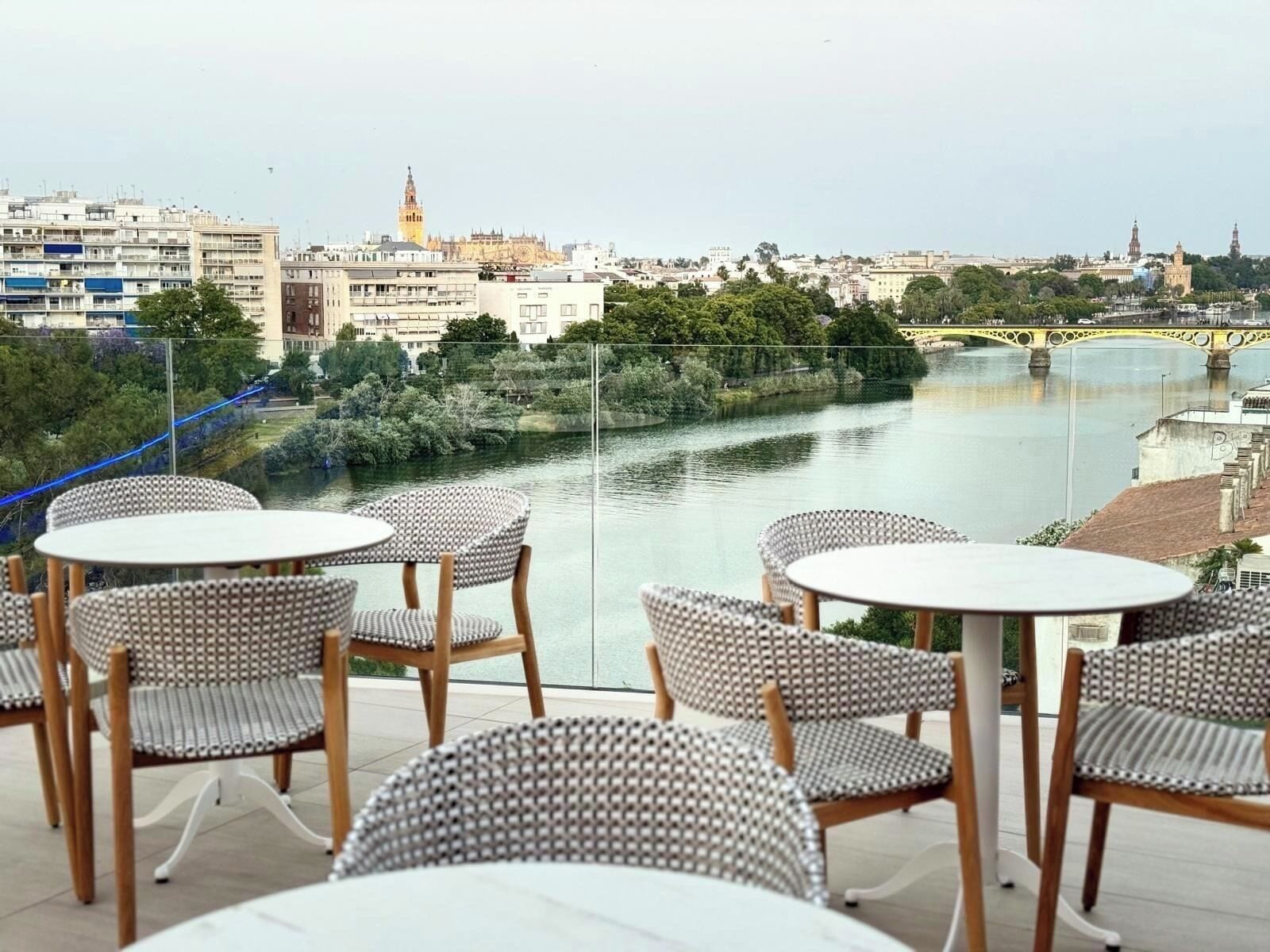 Um terraço com cadeiras e mesas de vime com vista para um rio, com edifícios e uma ponte ao fundo.