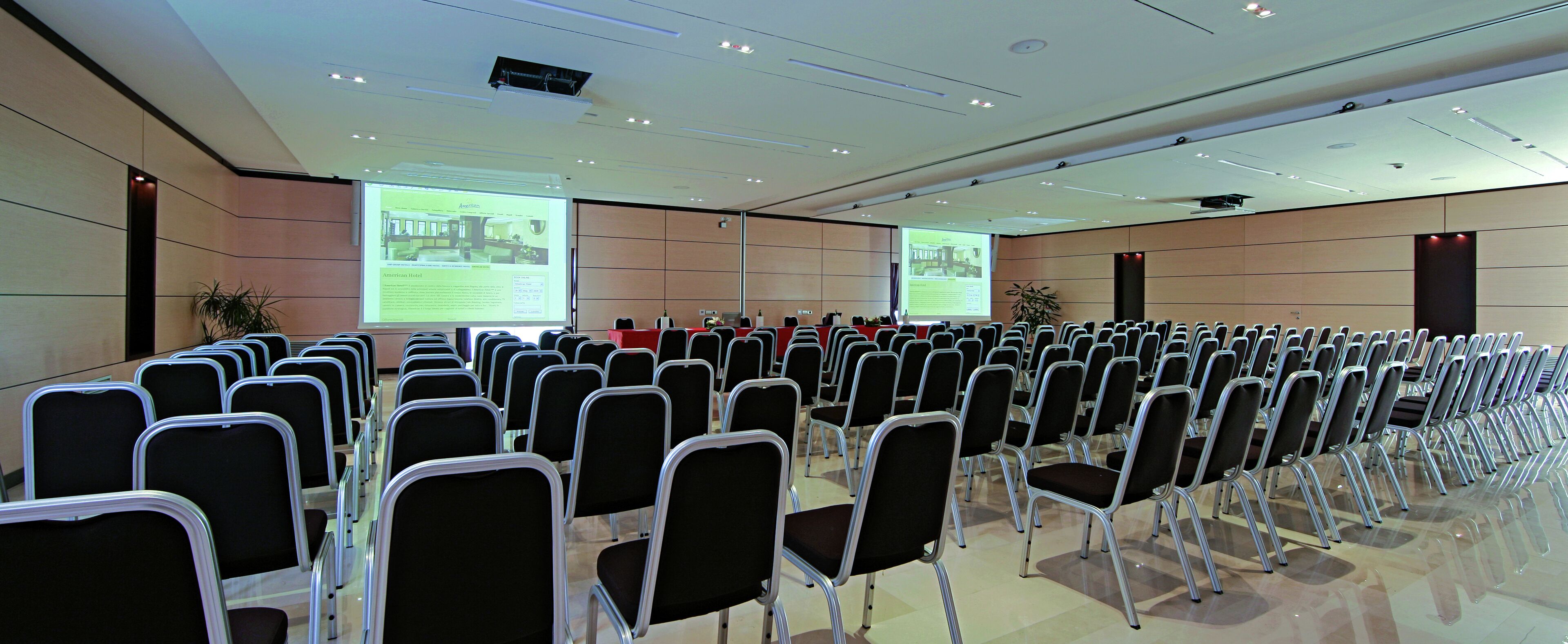 Uma sala de conferências com fileiras de cadeiras, um púlpito e duas telas de projeção exibindo o site de um hotel.
