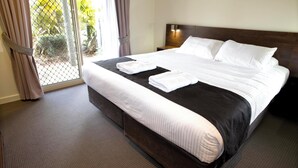 Quarto de hotel com cama grande, cabeceira com luminária e vista para vegetação através de uma janela com cortinas.