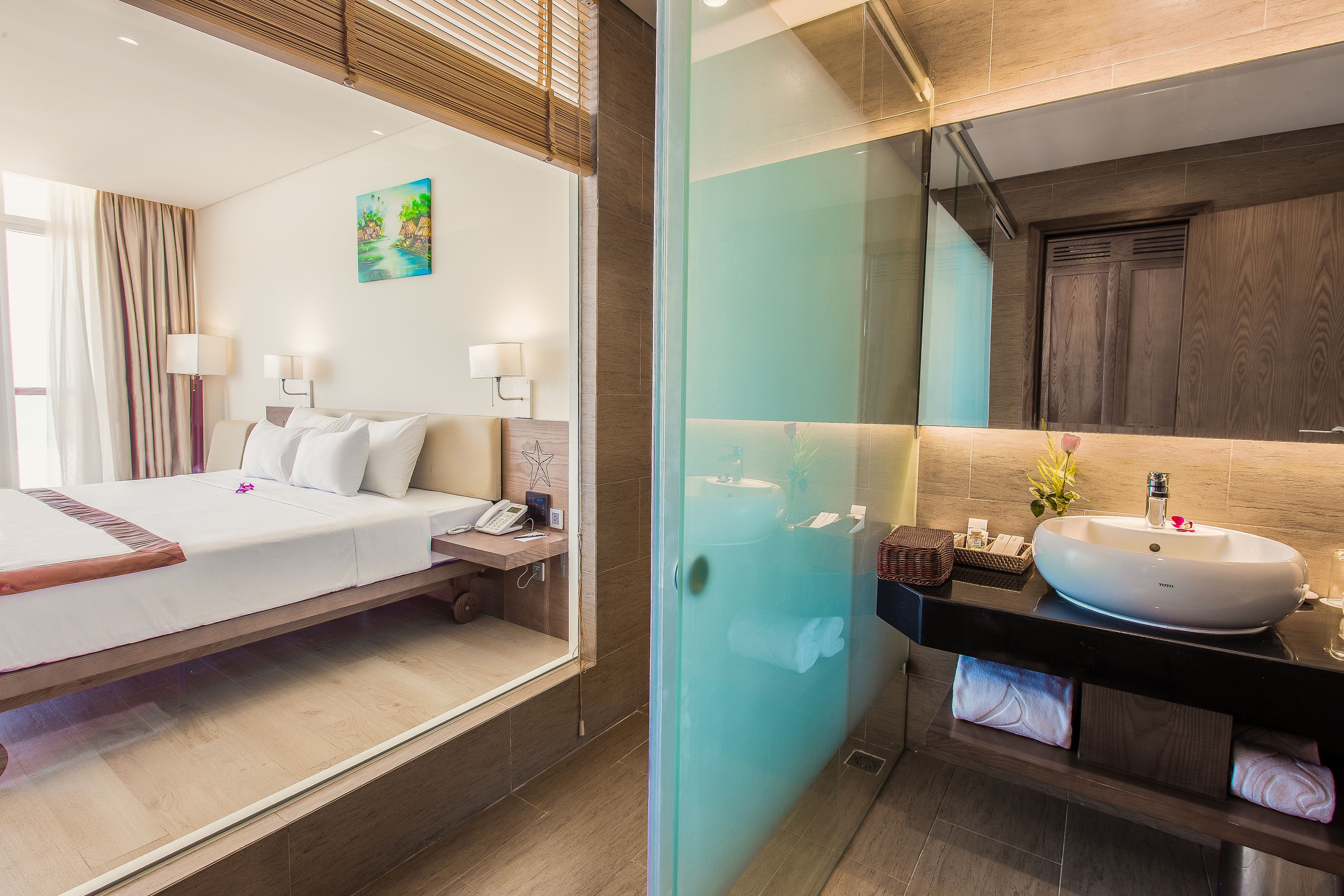 Quarto de hotel moderno com uma cama grande, uma mesa de cabeceira com abajur, um criado-mudo com telefone e um banheiro com pia e espelho espaçosos.