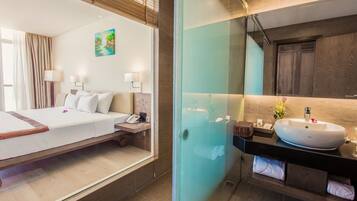 Quarto de hotel moderno com uma cama grande, uma mesa de cabeceira com abajur, um criado-mudo com telefone e um banheiro com pia e espelho espaçosos.