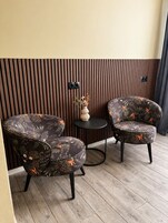 Quarto com duas poltronas, uma mesa pequena, parede de ripas de madeira e um vaso com flores secas.