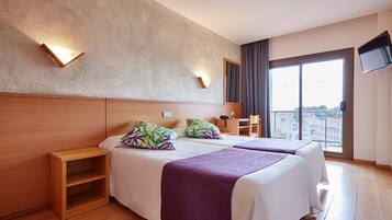 Quarto de hotel com duas camas, cabeceira de madeira, mesa de cabeceira, televisão e uma janela grande com vista para a cidade.