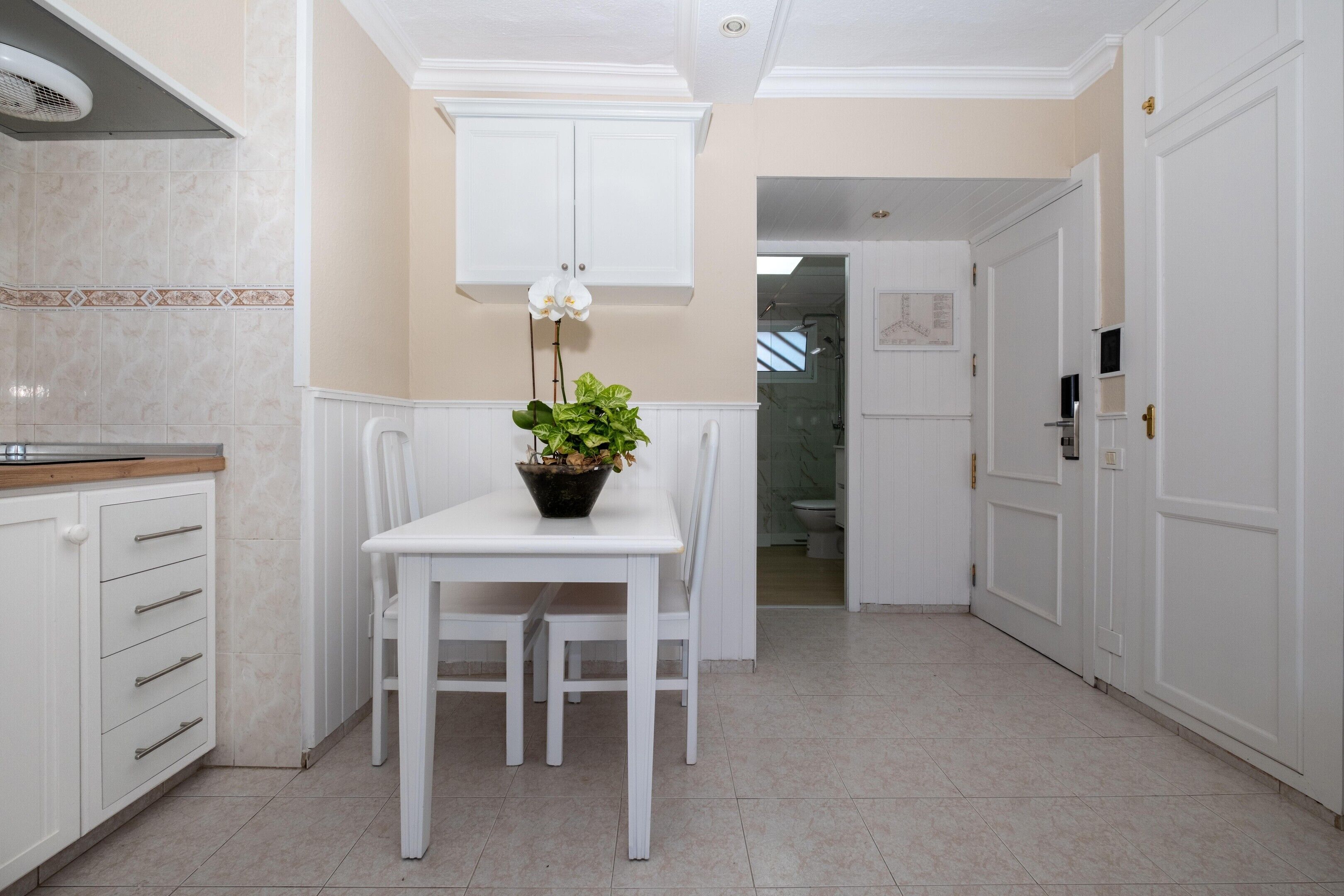 Uma cozinha com armários brancos, uma mesa de jantar branca com cadeiras, uma planta em vaso e um ventilador de teto branco.