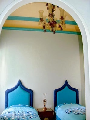 Quarto com duas camas, cada uma com cabeceira azul, um lustre e uma pequena mesa com abajur.