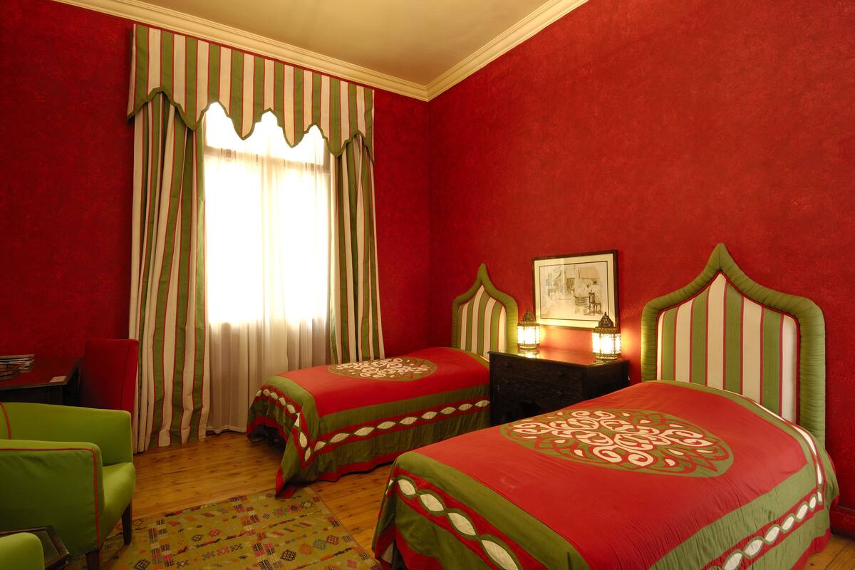 Quarto com duas camas, uma poltrona verde, piso de madeira e uma janela com cortinas listradas.