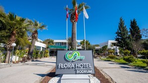 Placa do Hotel Flora Bodrum, com vista clara para a entrada do hotel, palmeiras e um edifício moderno ao fundo.