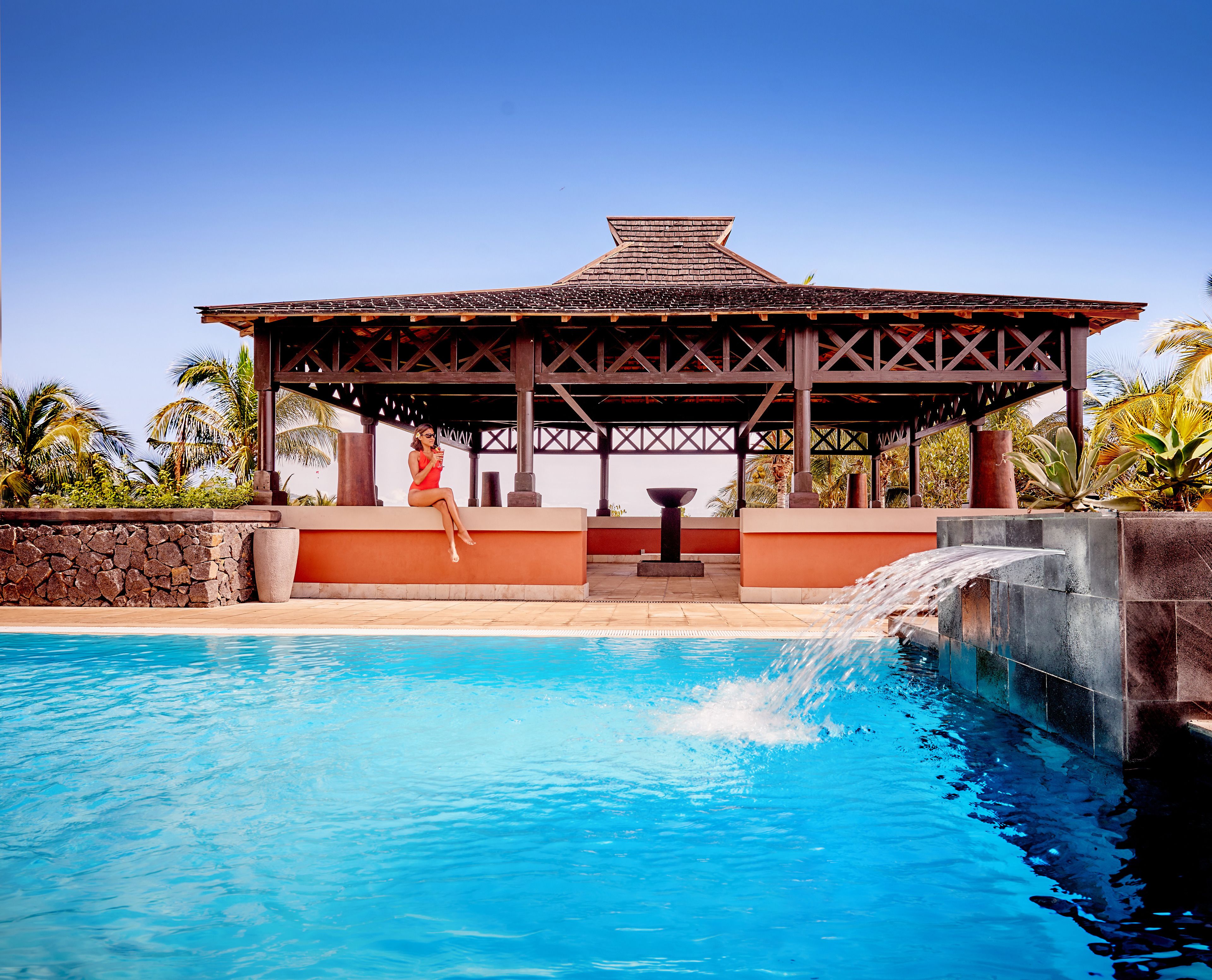 Uma área de piscina com um gazebo, uma parede de pedra e um elemento aquático.