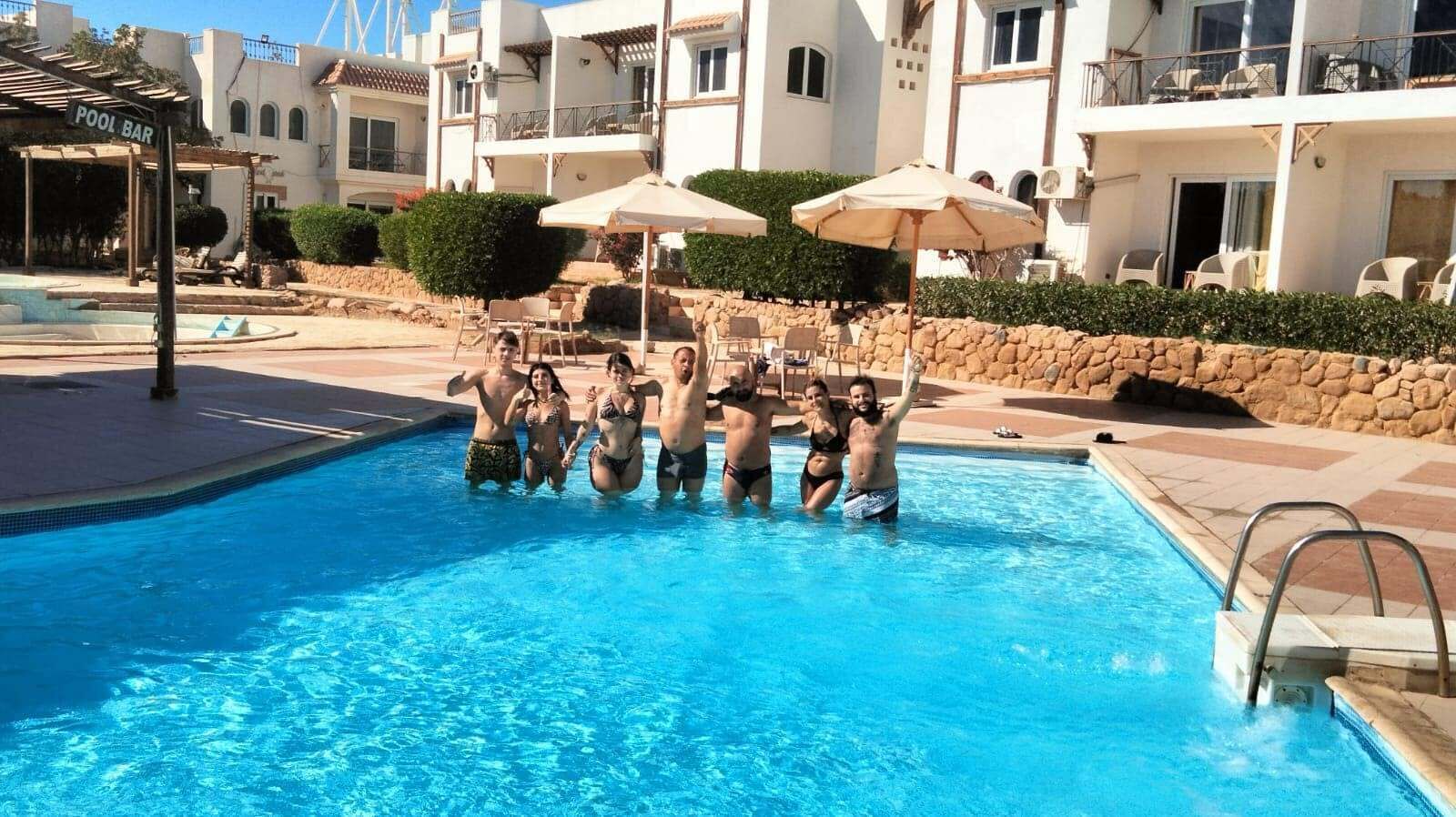 Piscina com pessoas na água, guarda-sóis e placa da piscina-bar.