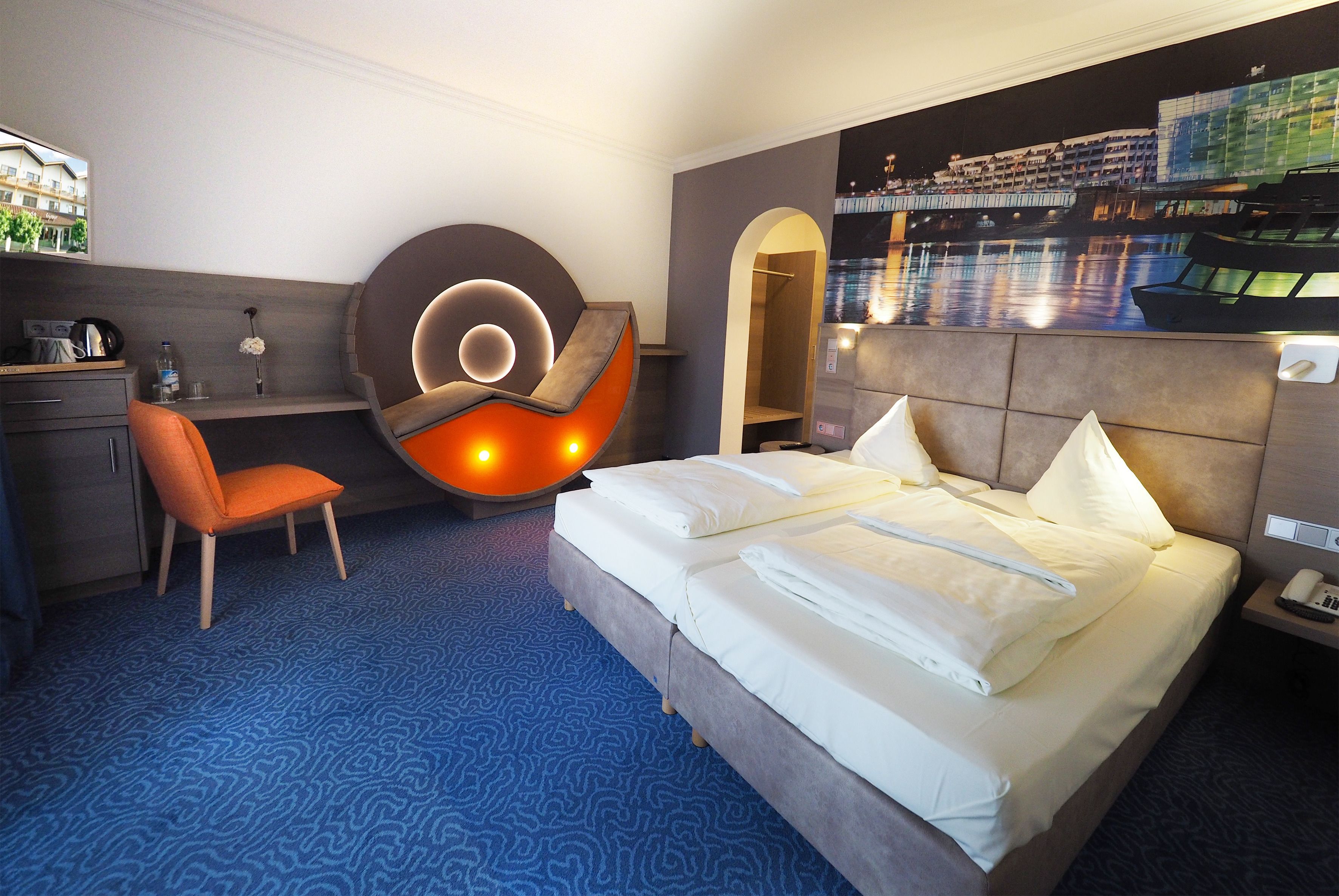 Quarto de hotel com uma cama, uma escrivaninha, uma cadeira e um grande painel decorativo na parede.