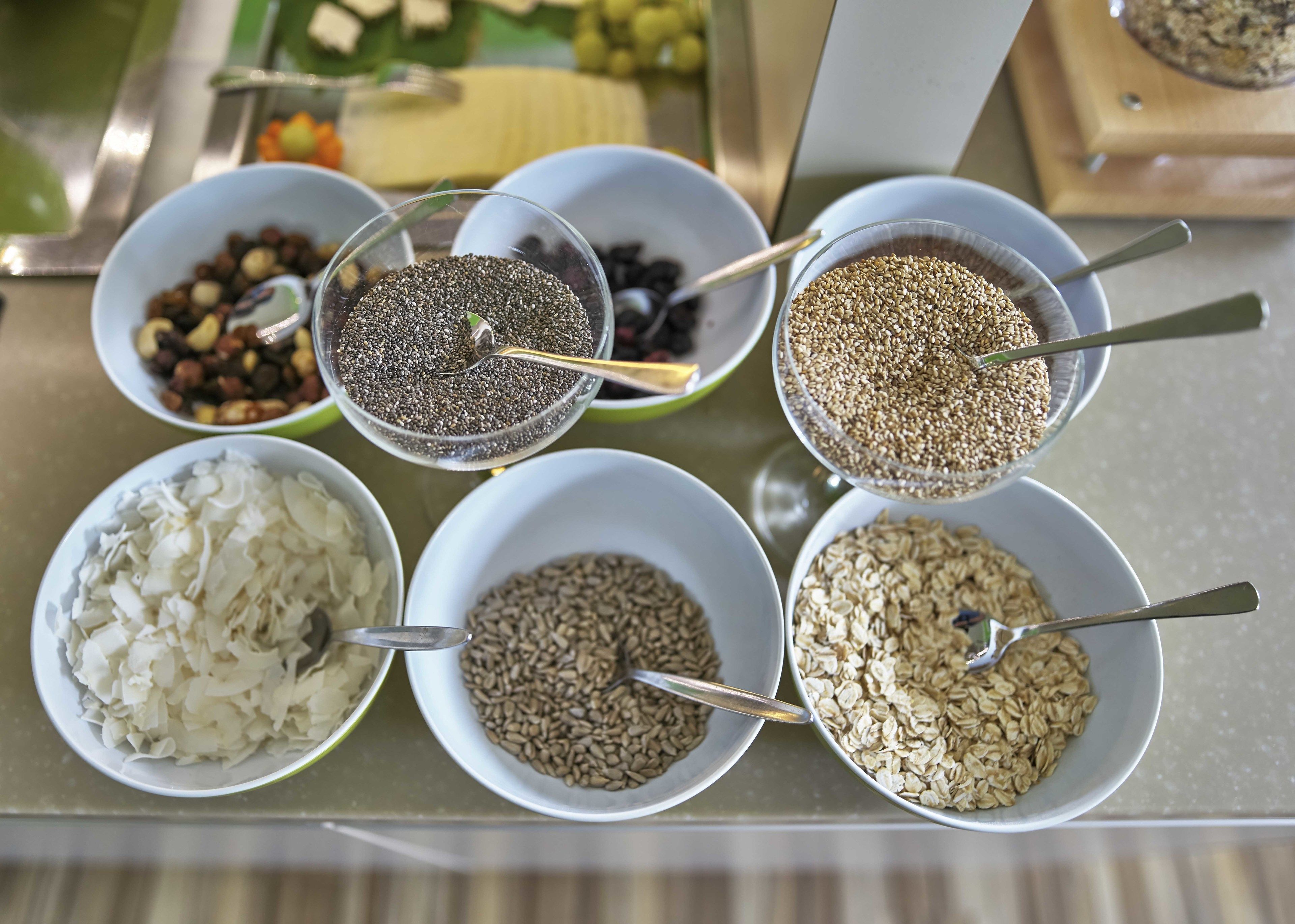 Diversos alimentos em recipientes, como nozes, sementes de chia e aveia, dispostos sobre uma mesa.
