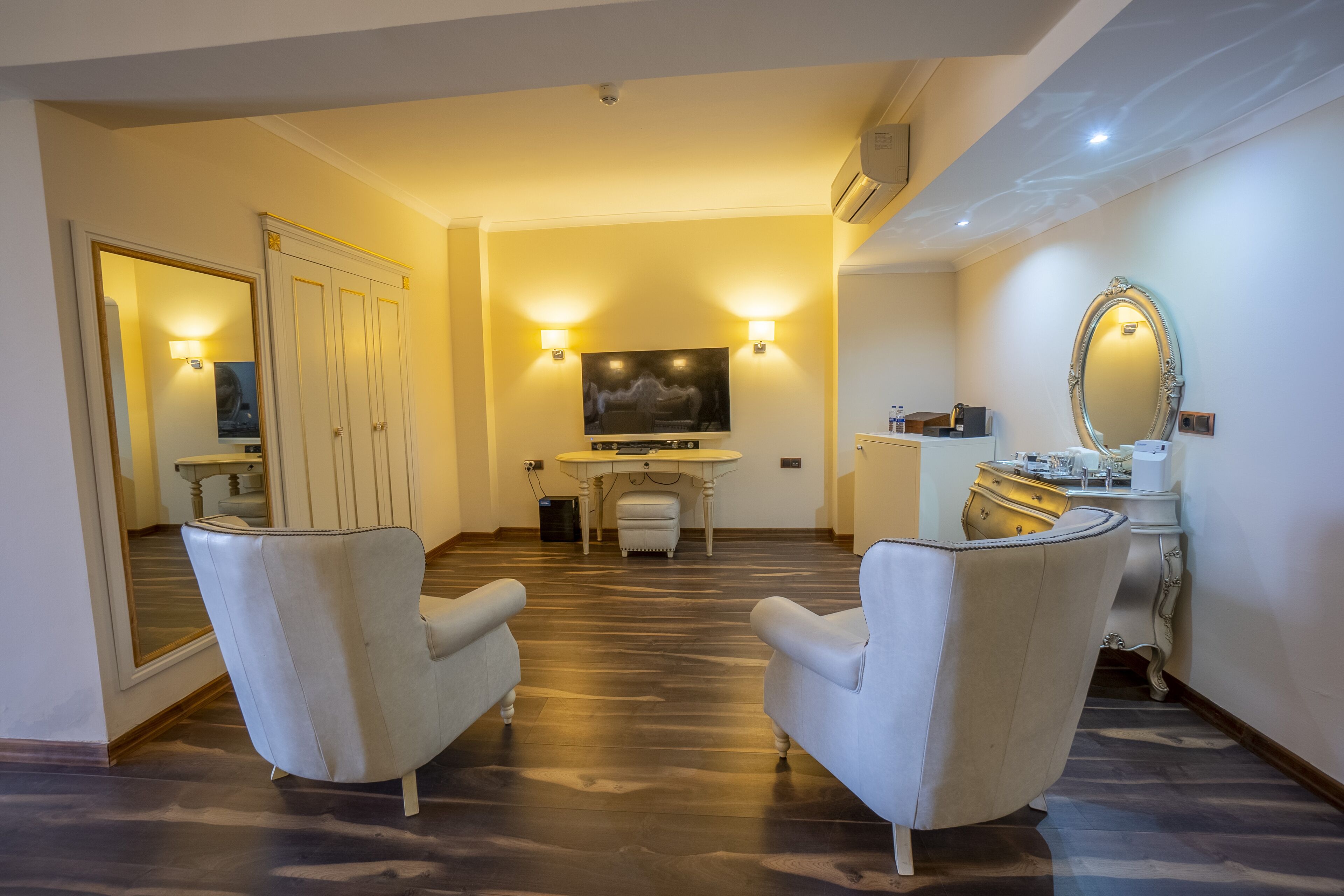 Quarto de hotel com um espelho grande, duas poltronas, uma toucador com espelho, uma televisão e luminárias fixadas na parede.