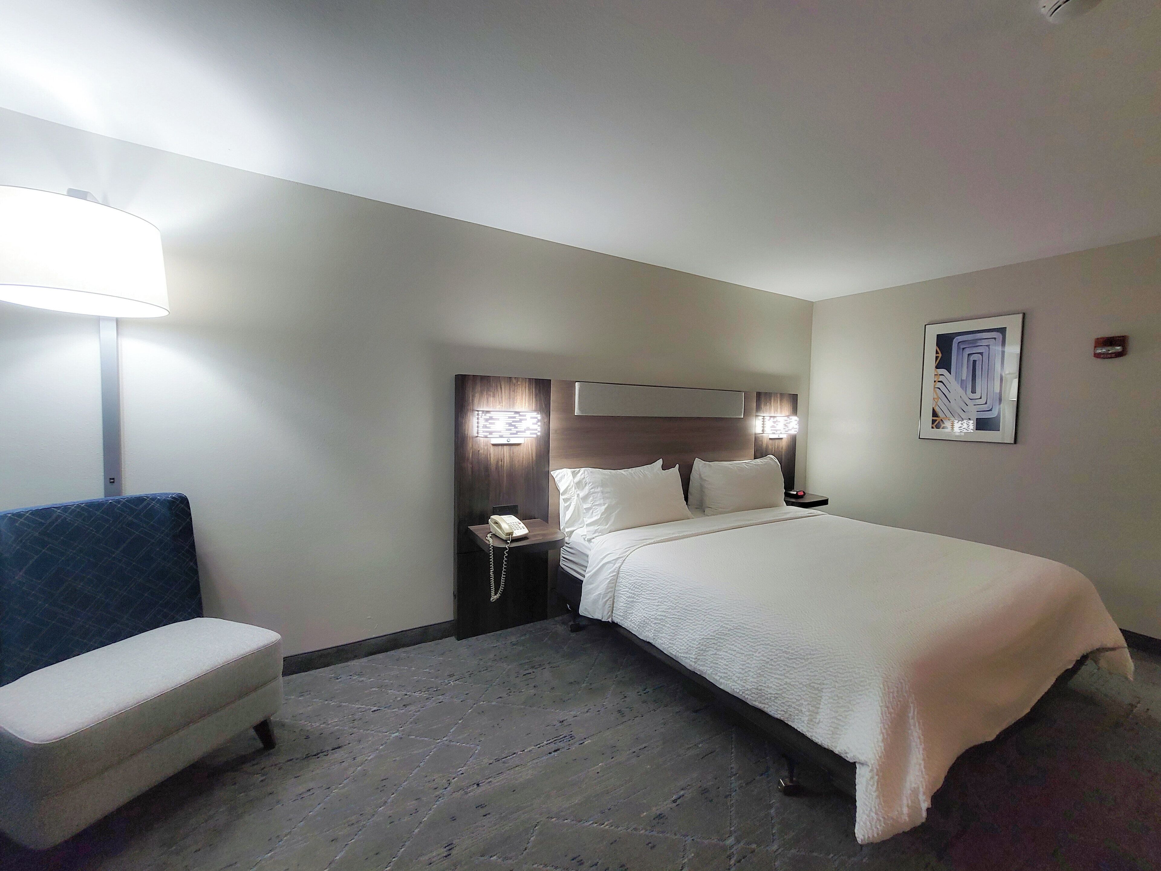 Quarto de hotel com cama, mesa de cabeceira, luminária e uma cadeira.