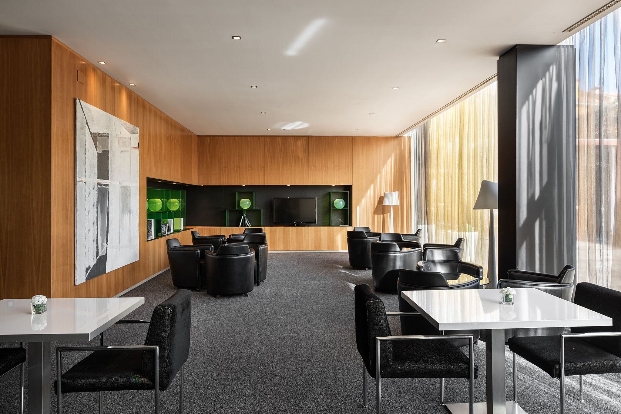 Um hall de hotel moderno com lambris de madeira, uma grande TV de tela plana e uma área de estar com cadeiras e mesas pretas.