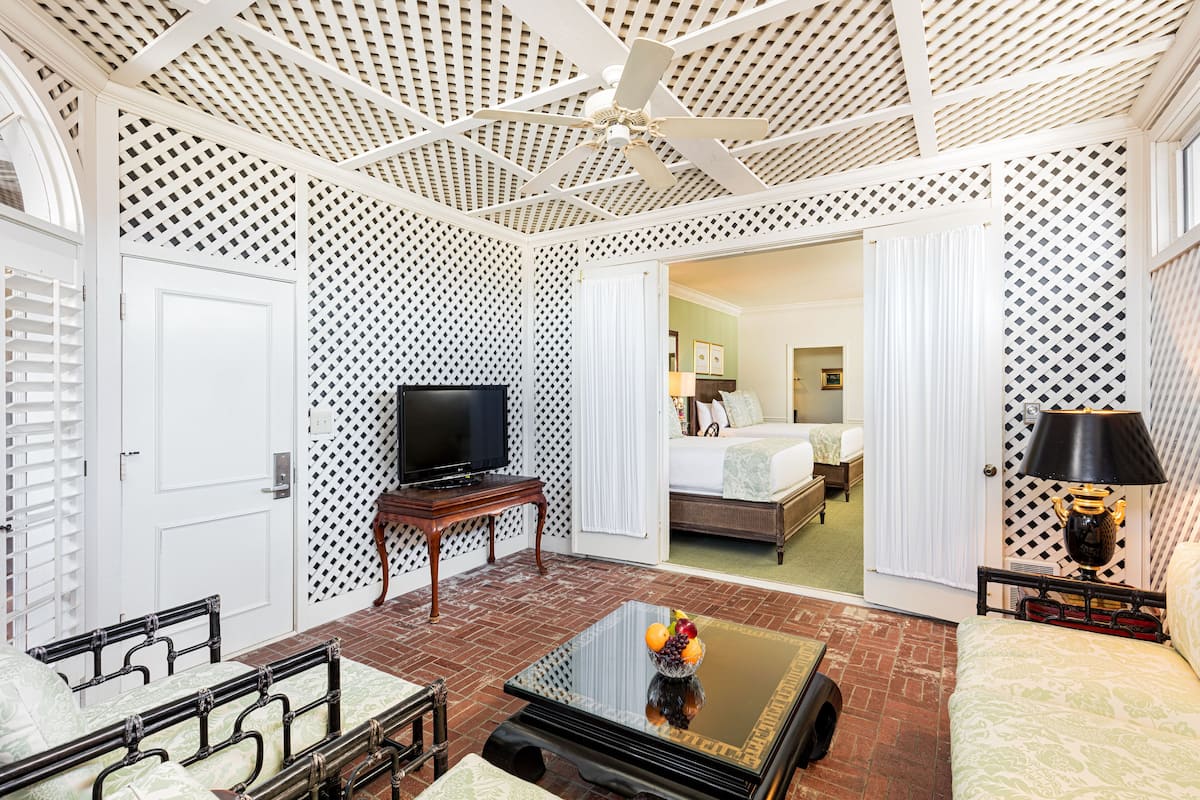Quarto de hotel com cama, televisão, mesinha de centro com frutas e ventilador de teto.