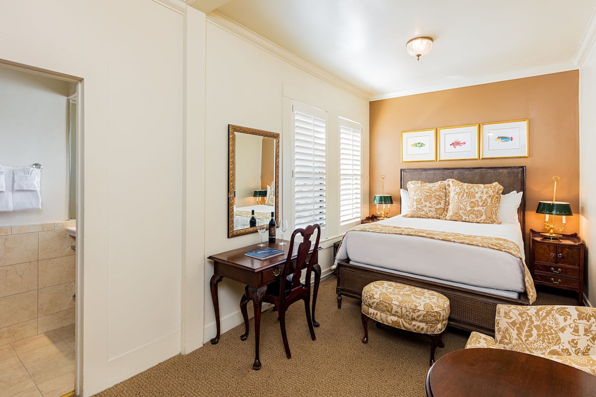 Quarto de hotel com uma cama, escrivaninha, cadeira, mesa de cabeceira e abajur.