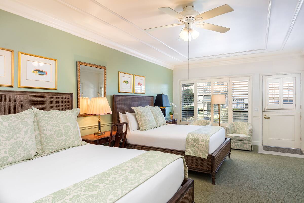 Quarto de hotel com duas camas, um ventilador de teto, um espelho grande e vista para o exterior pela janela.