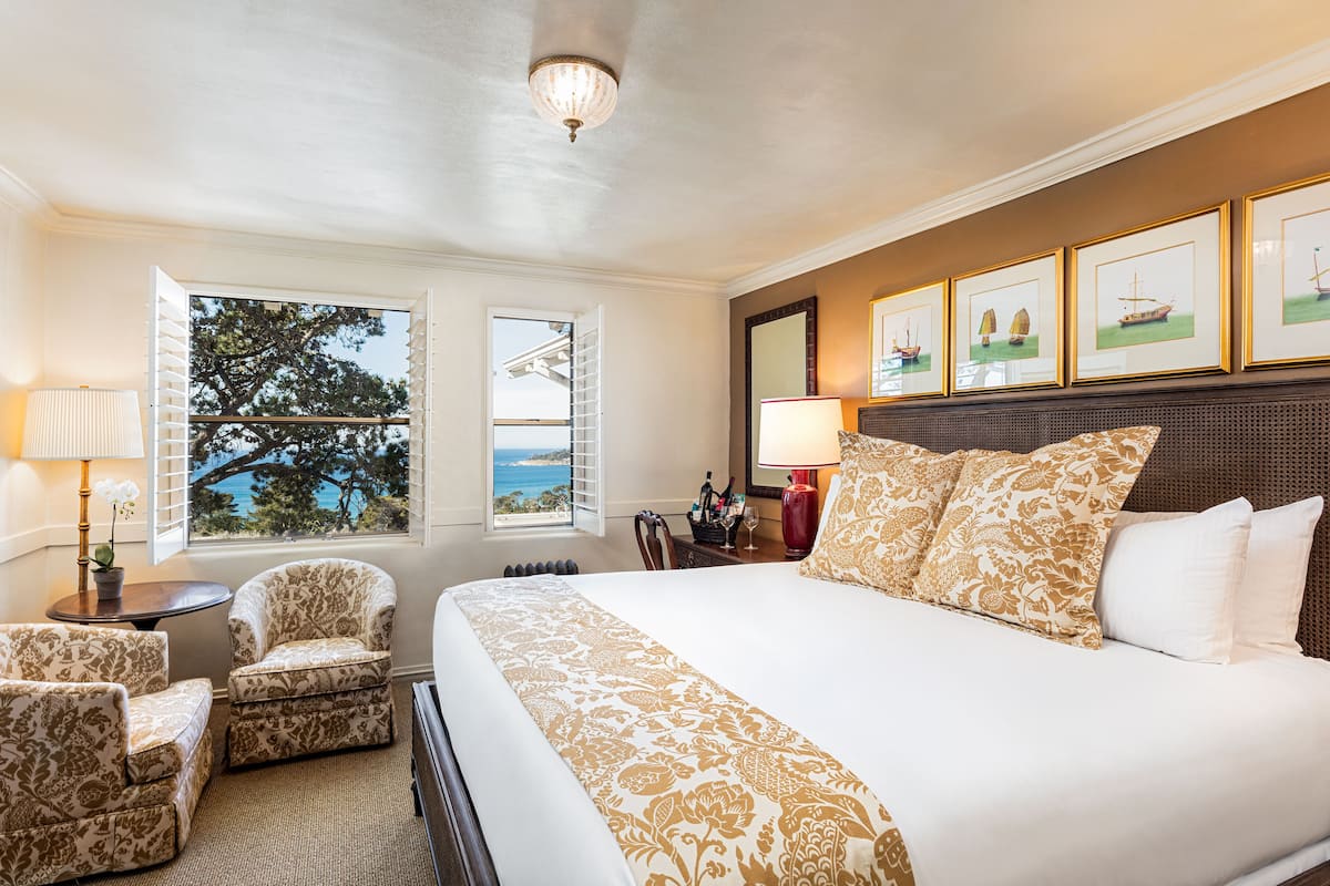 Quarto de hotel com uma cama grande, duas poltronas, uma mesinha e uma janela com vista para o oceano.