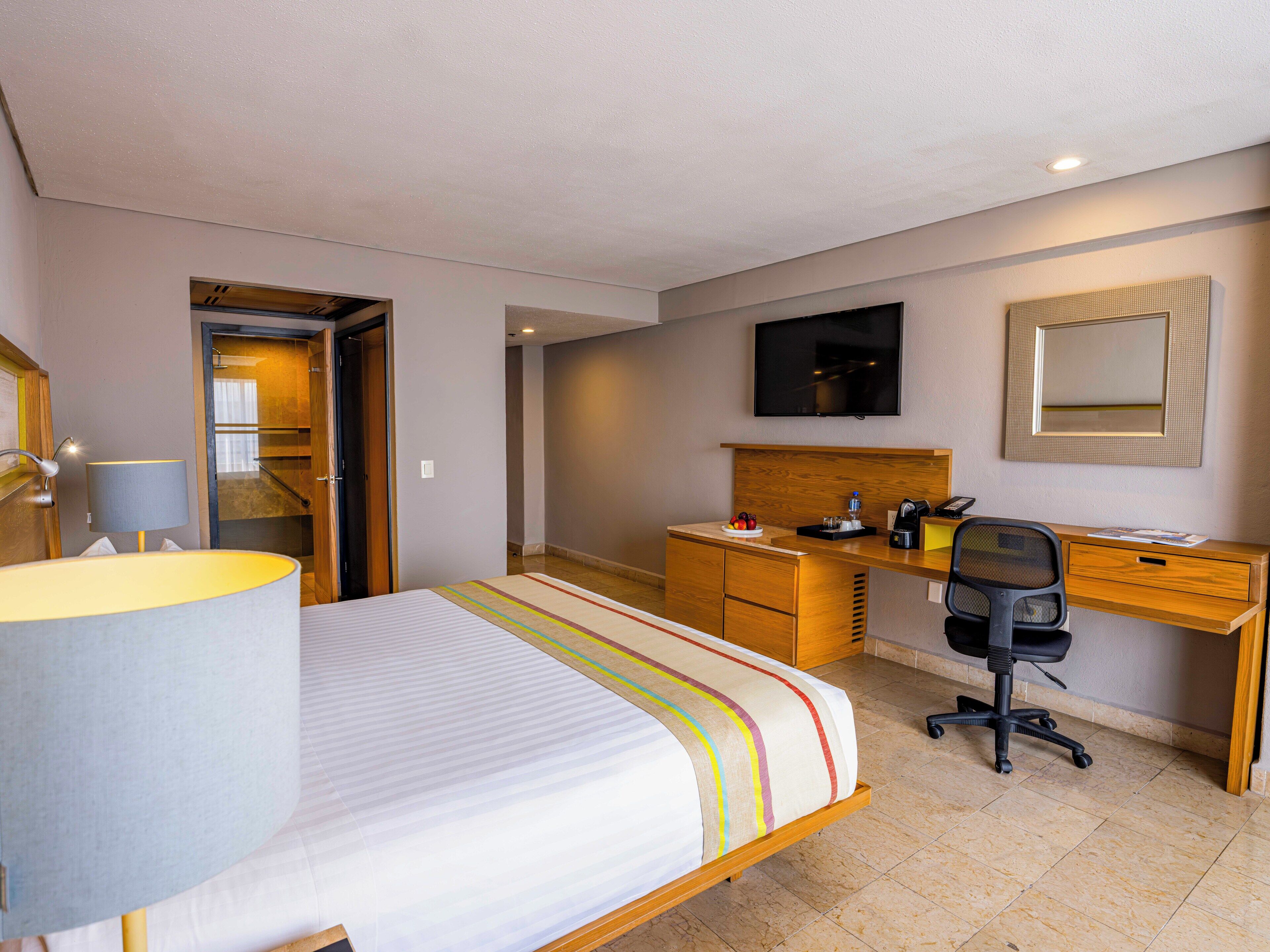 Quarto de hotel com cama, escrivaninha, cadeira e televisão na parede.