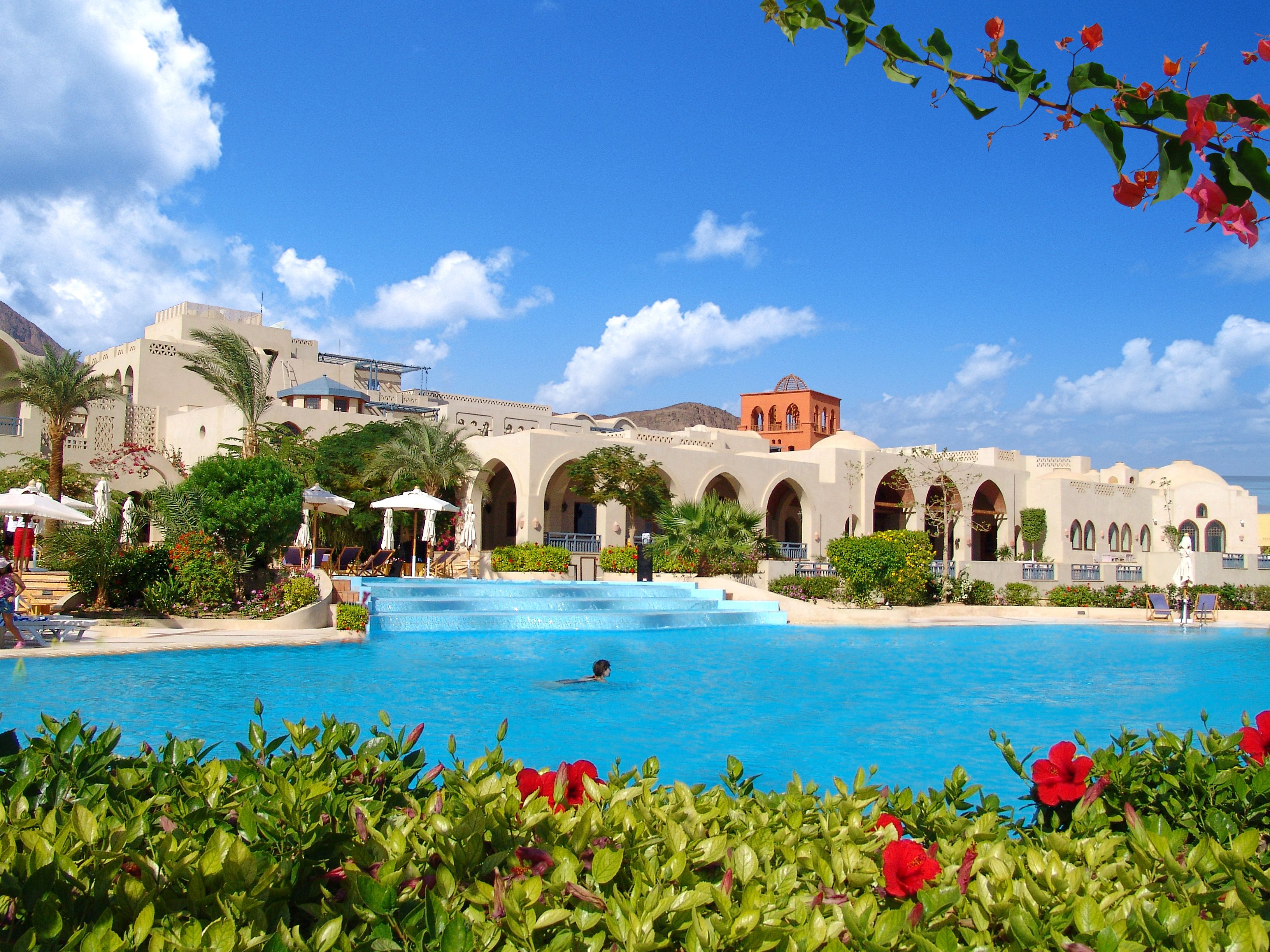 Piscina de resort com águas azuis cristalinas, rodeada por vegetação exuberante e flores vermelhas.