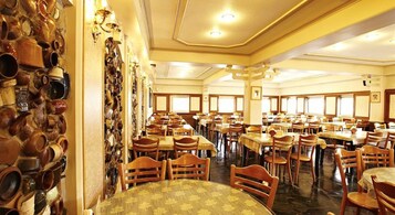 Uma ampla sala de jantar com várias mesas e cadeiras, uma parede decorativa com diversos objetos e um teto alto com iluminação embutida.