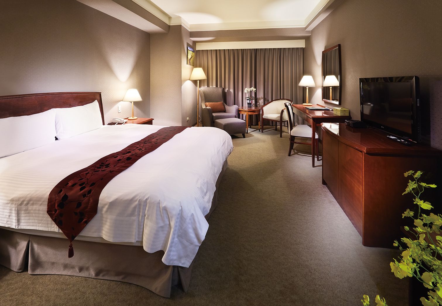 Quarto de hotel com uma cama grande, uma escrivaninha com televisão, uma cadeira e uma mesa pequena.