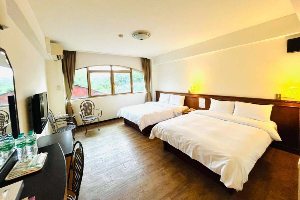Quarto de hotel com duas camas, uma escrivaninha com cadeira, televisão e janela com cortinas.