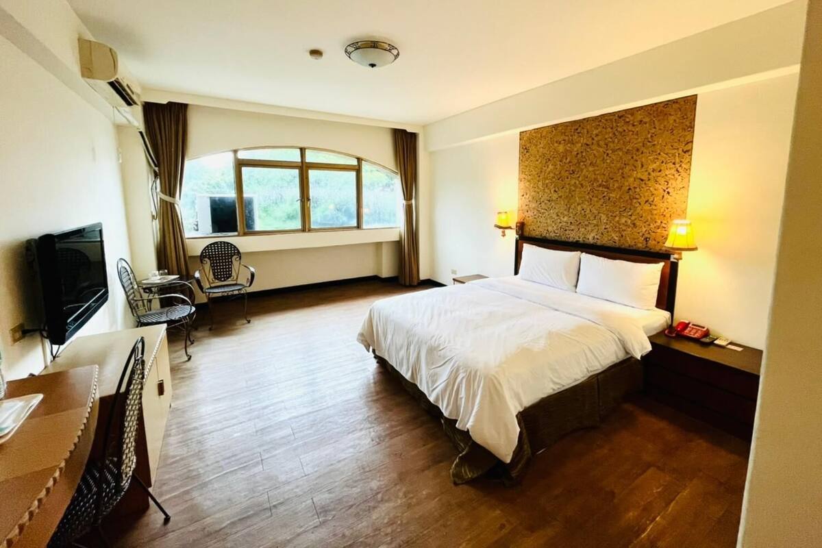 Quarto de hotel com uma cama grande, televisão, escrivaninha com cadeira, janela com cortinas e luminária na parede.