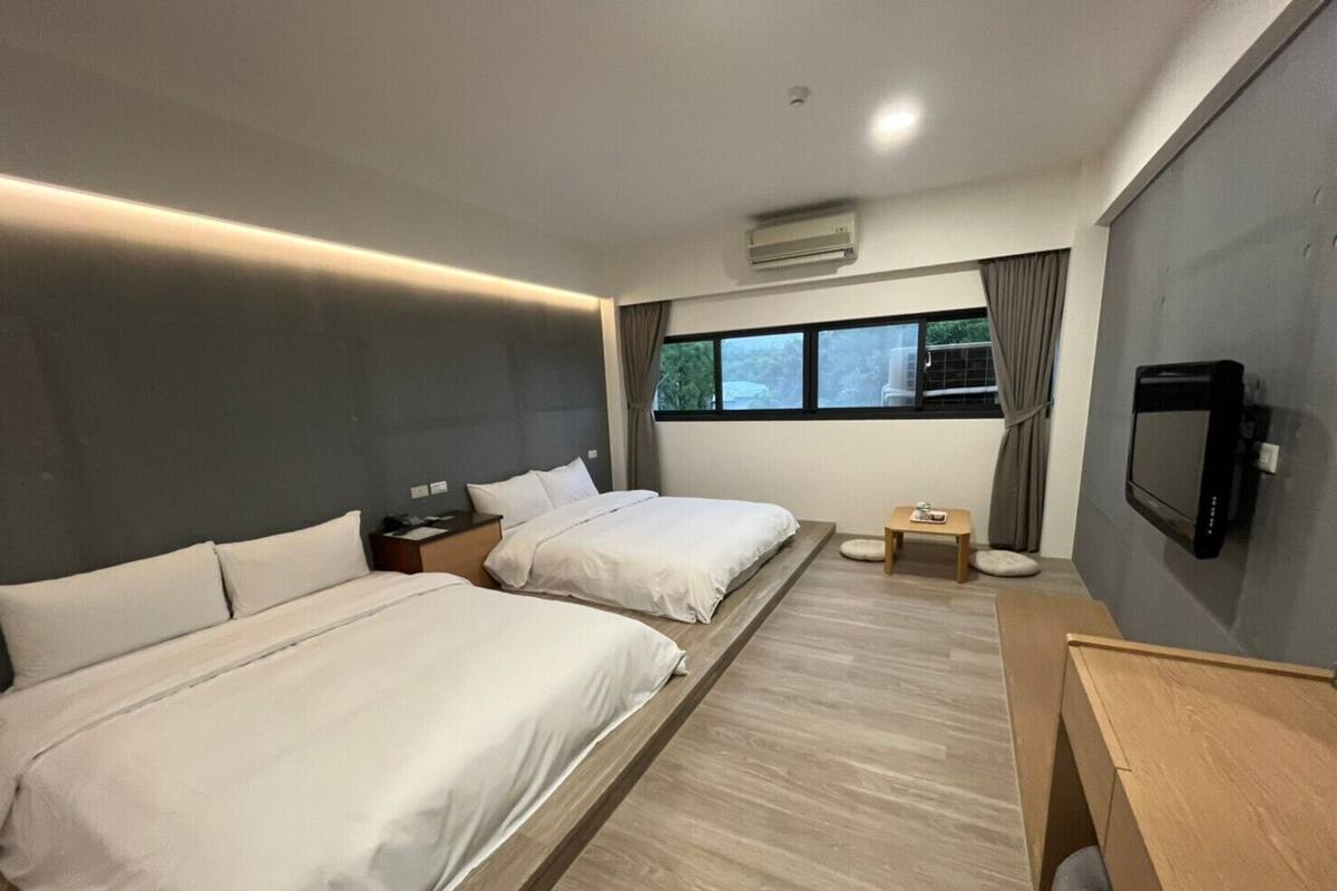 Quarto de hotel moderno com duas camas, uma mesa de cabeceira pequena, uma televisão e uma janela com cortinas.
