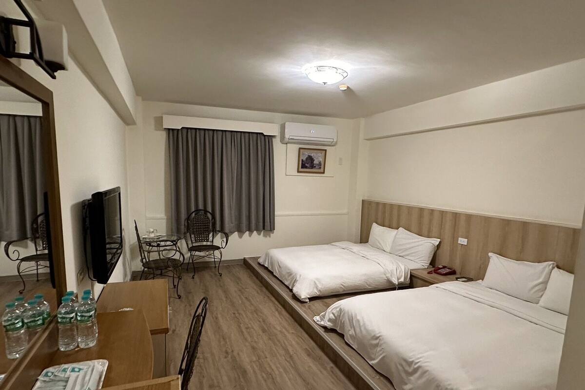 Quarto de hotel com duas camas, uma escrivaninha com cadeira, televisão e janela com cortinas.