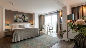 Quarto de hotel moderno com uma cama grande, uma escrivaninha e uma cadeira. Há um quadro na parede e um vaso com flores na escrivaninha.