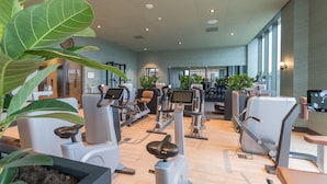 Academia moderna com diversos equipamentos de ginástica, grandes janelas e plantas dentro de casa.