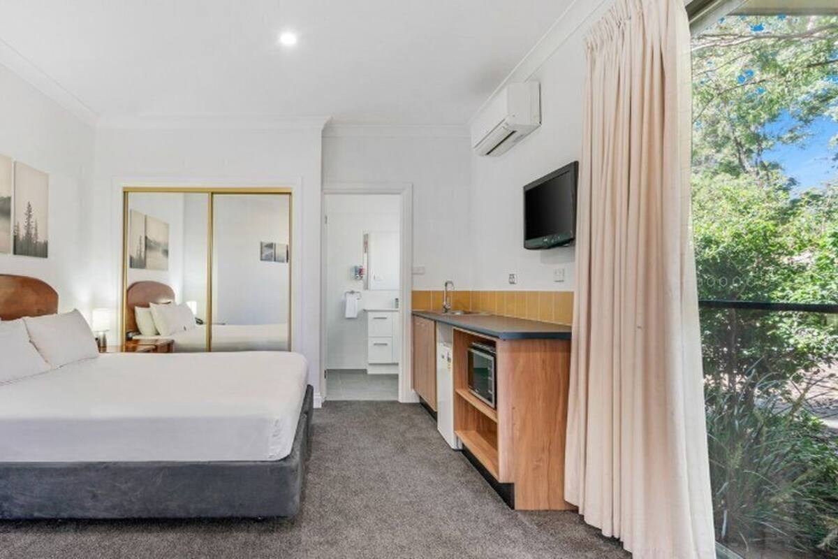 Quarto de hotel com cama, mesa de cabeceira, espelho, televisão, cozinha compacta e varanda com cortinas.