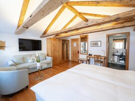 Quarto moderno com vigas de madeira aparentes, uma cama grande, um sofá, uma pequena mesa com um vaso e um banheiro visível através de uma porta aberta.
