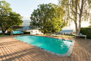 Piscina retangular com deck de tijolos, rodeada por árvores e um edifício branco com telhado azul.