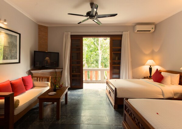 Quarto de hotel com cama, sofá, televisão, ventoinha no teto e varanda com vista para o verde.
