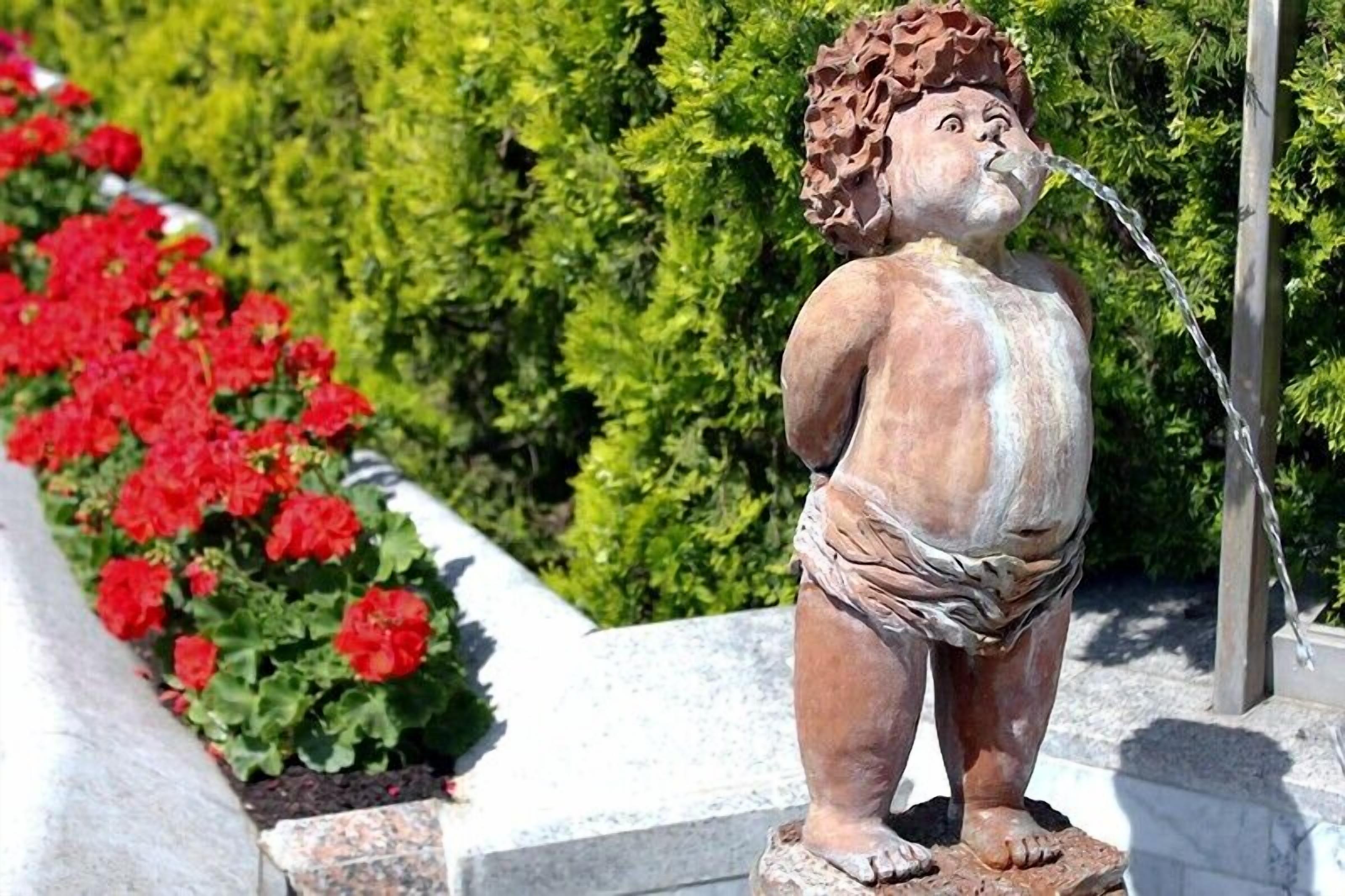 Estátua de uma criança bebendo água, rodeada por flores vermelhas e vegetação.