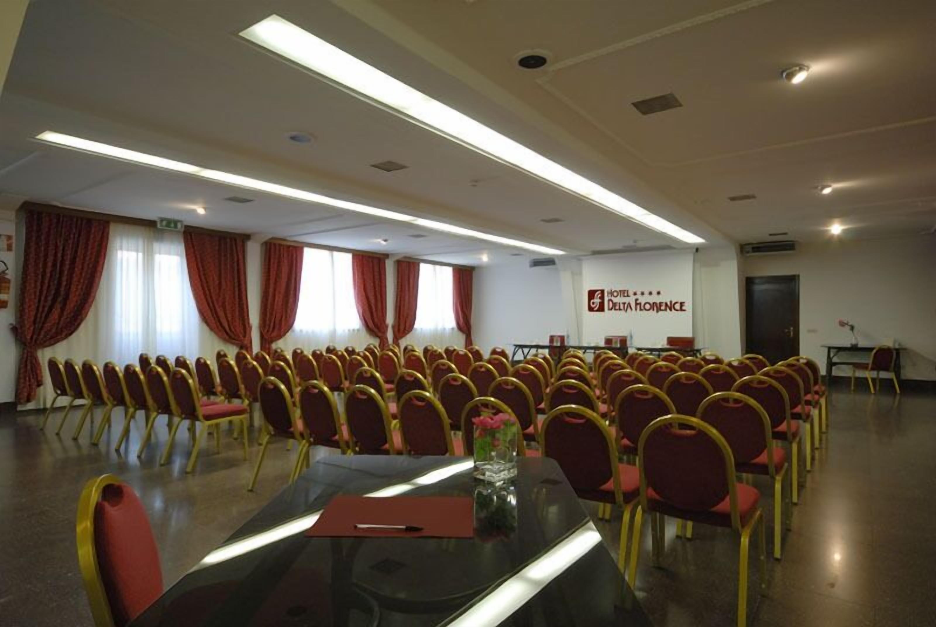 Uma sala de conferências com fileiras de cadeiras, uma mesa comprida com um vaso de flores e uma placa com o nome Hotel Delfia Florence.