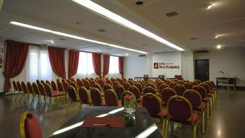 Uma sala de conferências com fileiras de cadeiras, uma mesa comprida com um vaso de flores e uma placa com o nome Hotel Delfia Florence.
