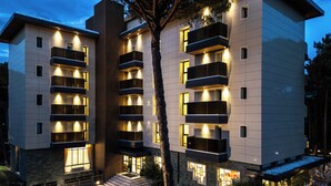 Um edifício moderno de vários andares com varandas, uma entrada bem iluminada e uma área externa ajardinada com assentos.